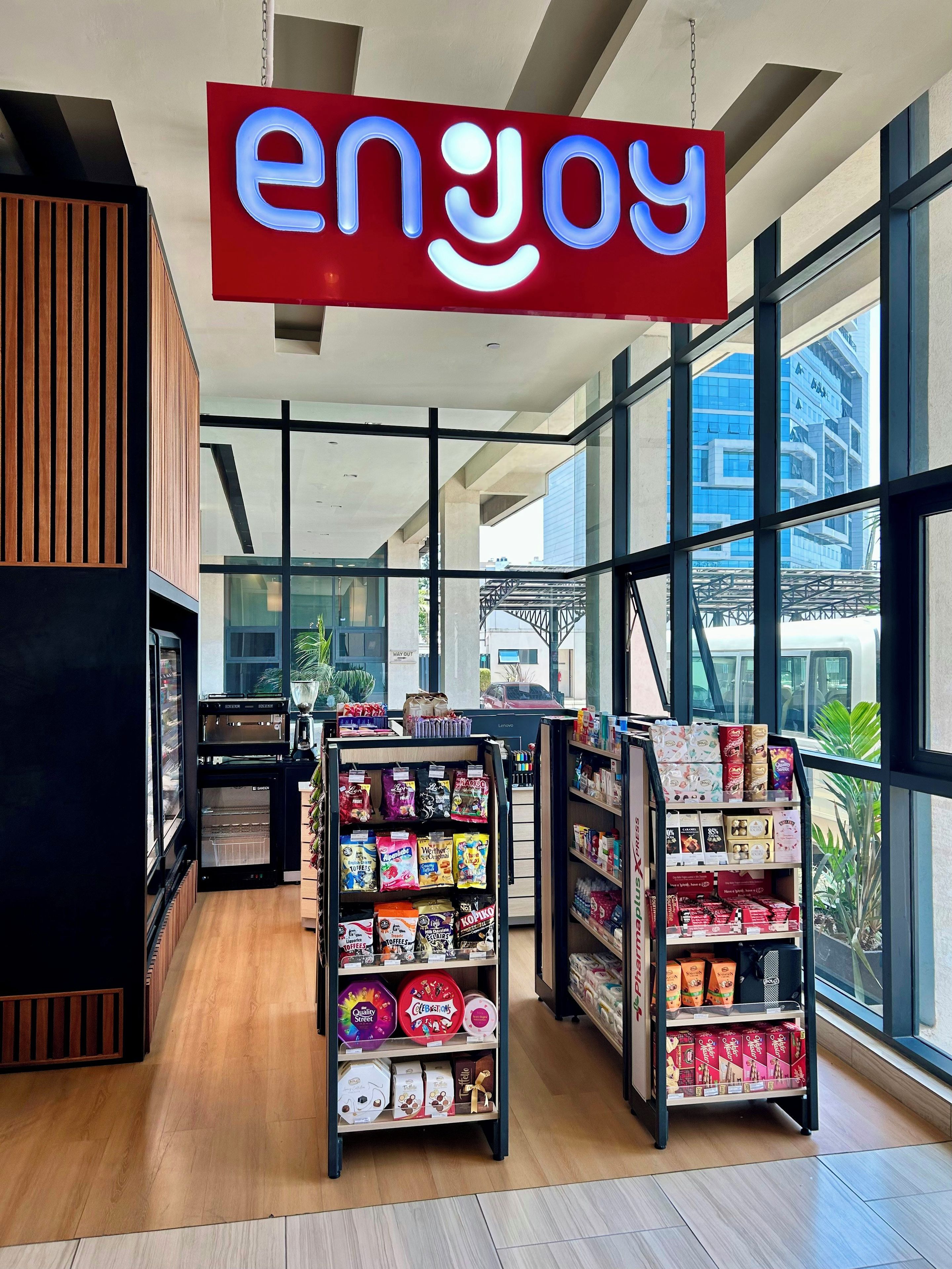 Interior de uma loja moderna, com um letreiro chamativo escrito 'enjoy', piso de madeira e prateleiras repletas de lanches e bebidas.
