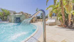 Piscina com cascata, cercada por espreguiçadeiras e plantas tropicais.