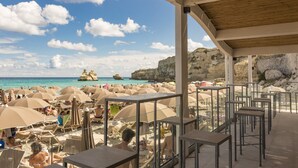 Terraço à beira-mar com guarda-sóis e assentos, com vista para uma praia cheia e falésias rochosas.
