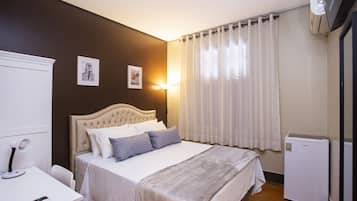 Quarto de hotel com cama, escrivaninha, luminária, controle remoto, aquecedor e janela com cortinas.