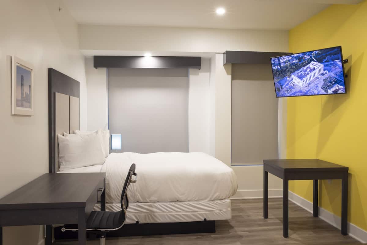 Quarto de hotel com cama, escrivaninha, cadeira, TV fixada na parede e uma pequena mesa.