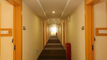 Corredor de hotel com portas em ambos os lados, piso coberto de carpete e extintor de incêndio vermelho.