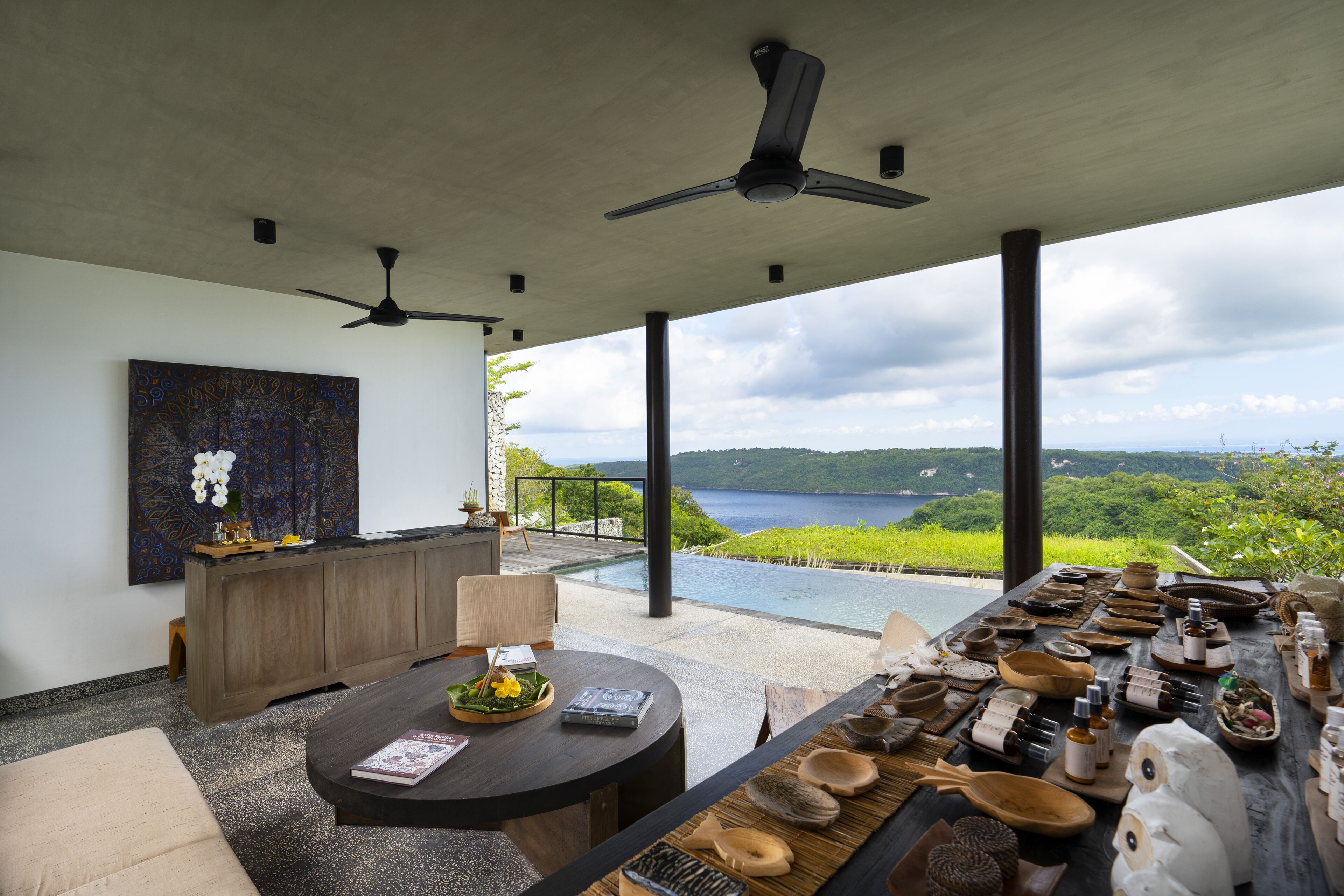 Uma sala de estar moderna com uma janela ampla que oferece vista para uma paisagem deslumbrante, um ventilador de teto e diversos objetos decorativos sobre a mesa de centro.