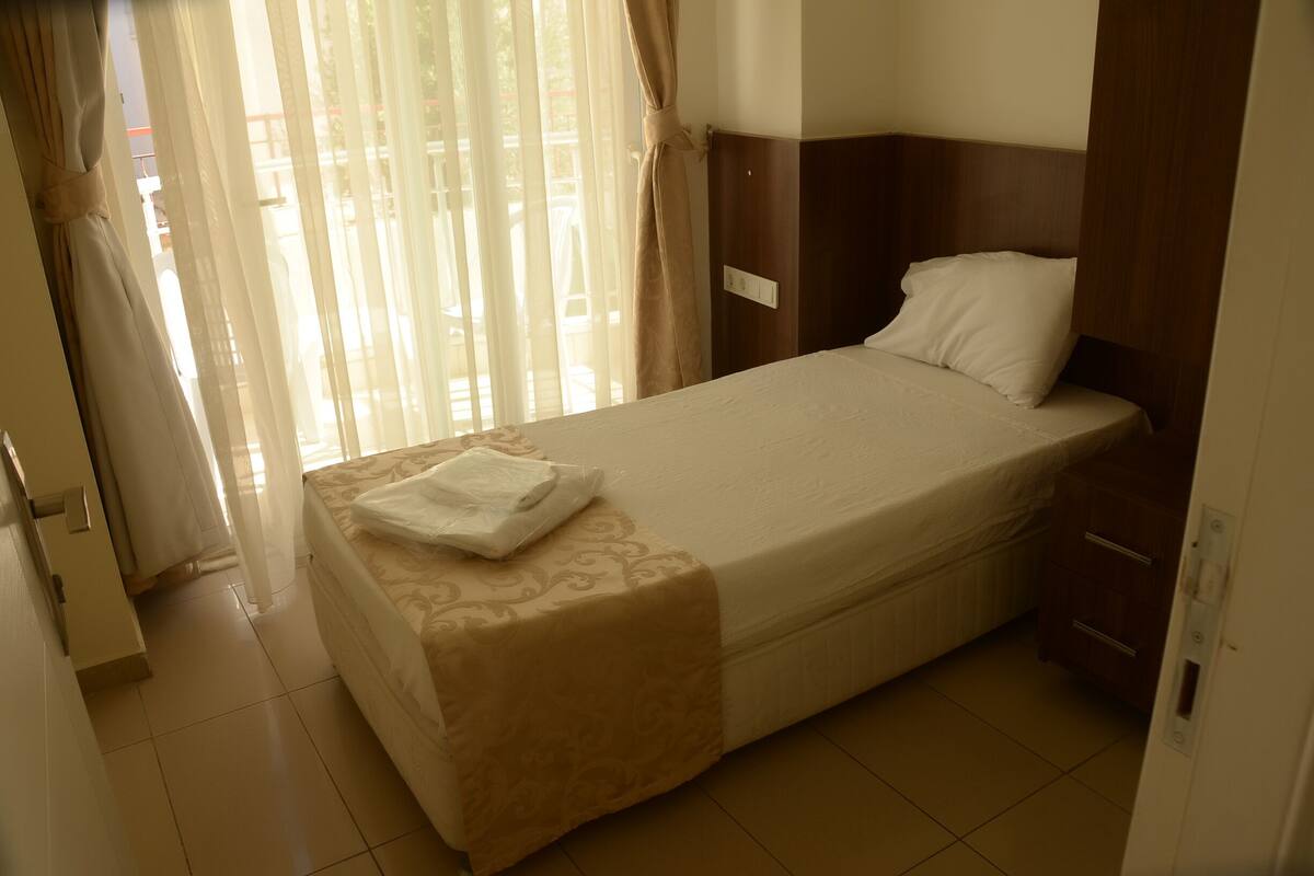 Uma cama de solteiro com um cobre-leito bege, um travesseiro branco e uma toalha branca dobrada. Há uma cabeceira de madeira e uma mesa de cabeceira de madeira à direita da cama.