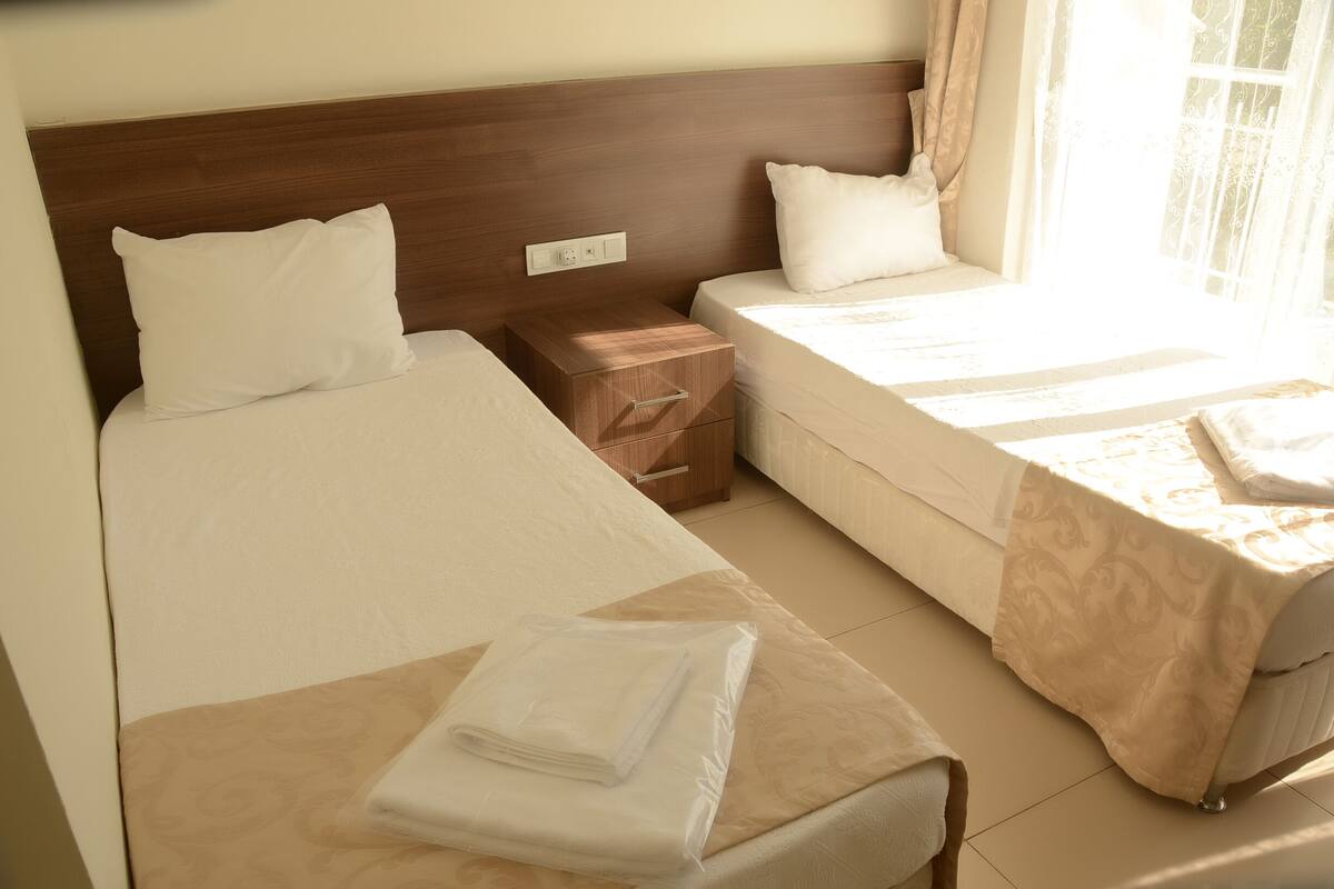Quarto de hotel com duas camas, cabeceira de madeira, mesa de cabeceira e janela com cortinas.
