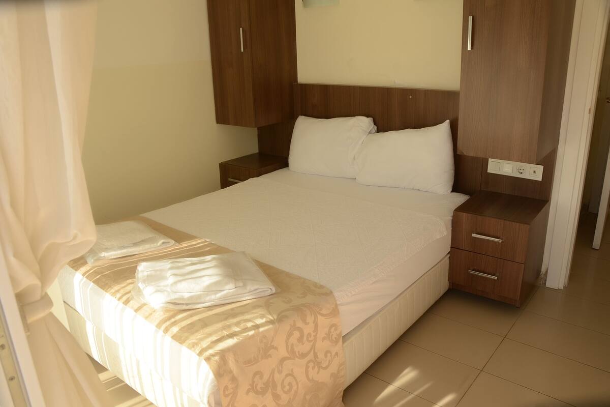 Cama bem arrumada com colcha estampada, mesa de cabeceira de madeira com gavetas e tomada elétrica na parede.