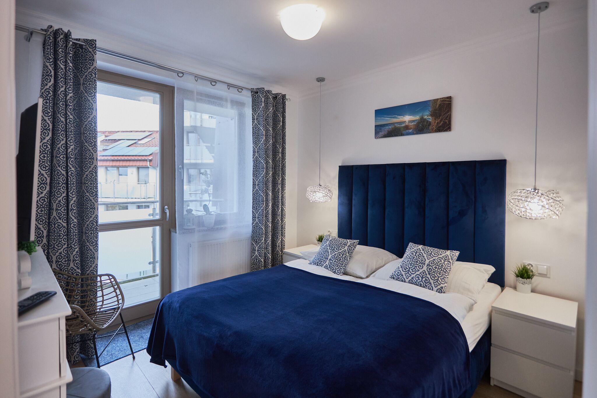 Quarto moderno com uma cama grande, vista da cidade através de uma porta deslizante e uma pintura de paisagem urbana na parede.
