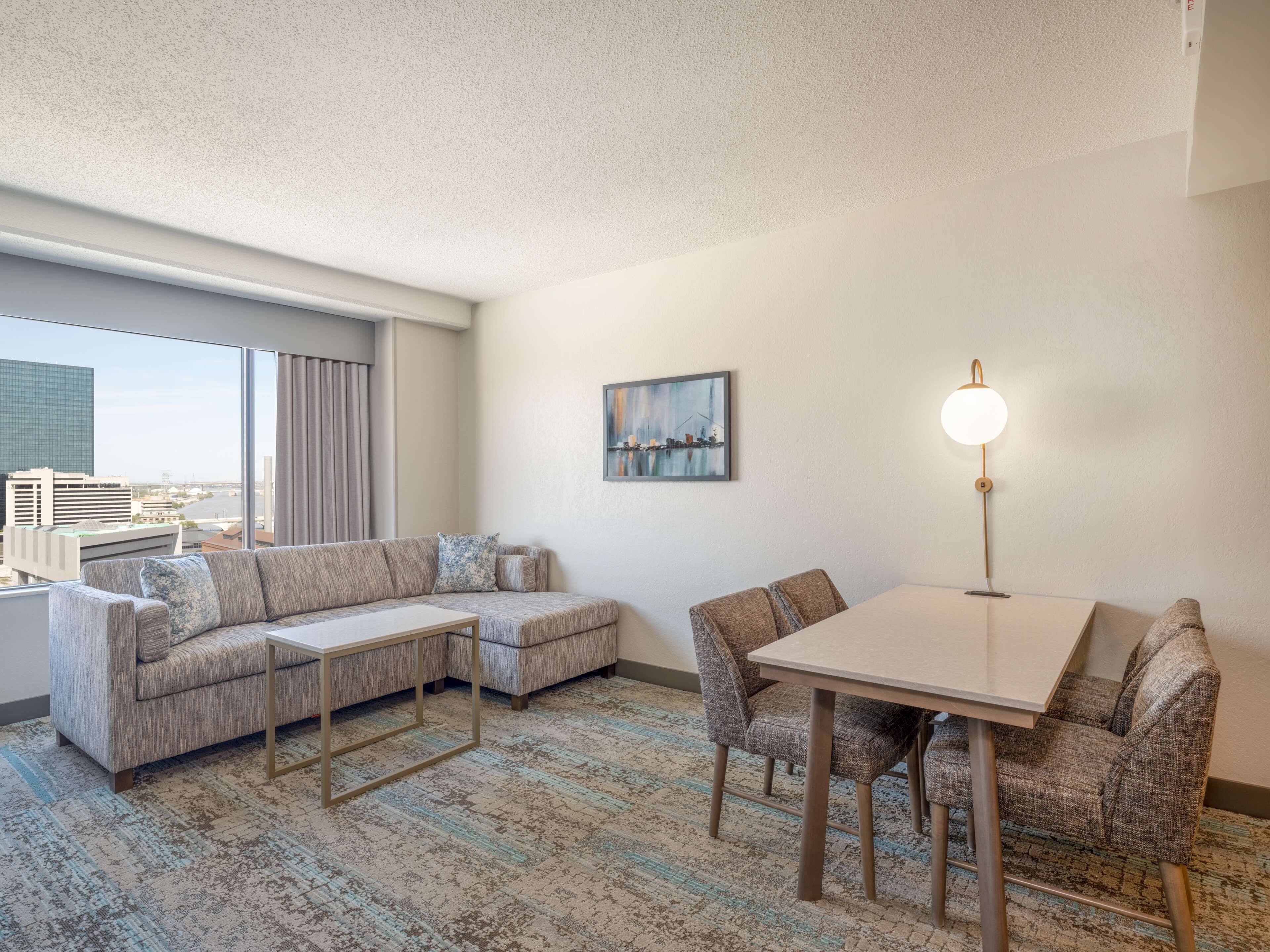 Quarto de hotel moderno com sofá modular, mesa de jantar com cadeiras, vista da cidade e um quadro na parede.