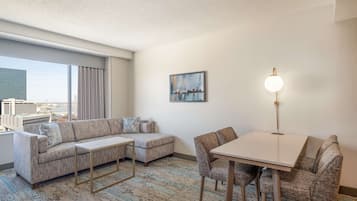 Quarto de hotel moderno com sofá modular, mesa de jantar com cadeiras, vista da cidade e um quadro na parede.
