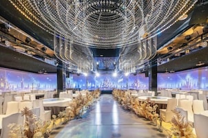 Uma sala de banquetes com fileiras de mesas e cadeiras brancas, uma passagem central e um teto decorado com luminárias pendentes.
