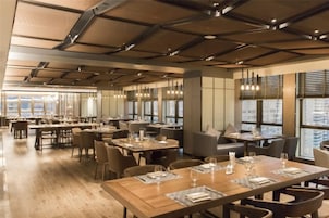 Um restaurante espaçoso com pé-direito alto, diversas mesas de jantar e amplas janelas que permitem a entrada de luz natural.