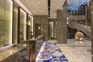 Um saguão de hotel com uma receção de mármore, uma escadaria e um candeeiro decorativo.