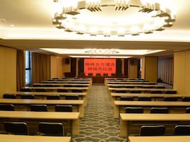 Uma sala de conferências com fileiras de mesas e cadeiras, uma tela grande na frente e um candelabro acima.