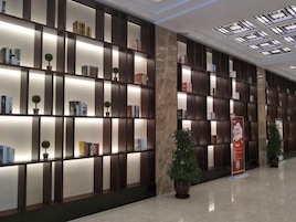 Um saguão de hotel moderno com um balcão de recepção extenso, piso de mármore e uma estante cheia de livros e plantas em vasos.
