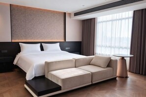 Quarto de hotel moderno com uma cama grande, um sofá, uma mesinha e uma janela ampla com cortinas.