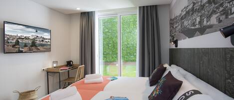 Quarto de hotel com cama, escrivaninha, cadeira, janela com cortinas e um quadro de uma vista urbana na parede.