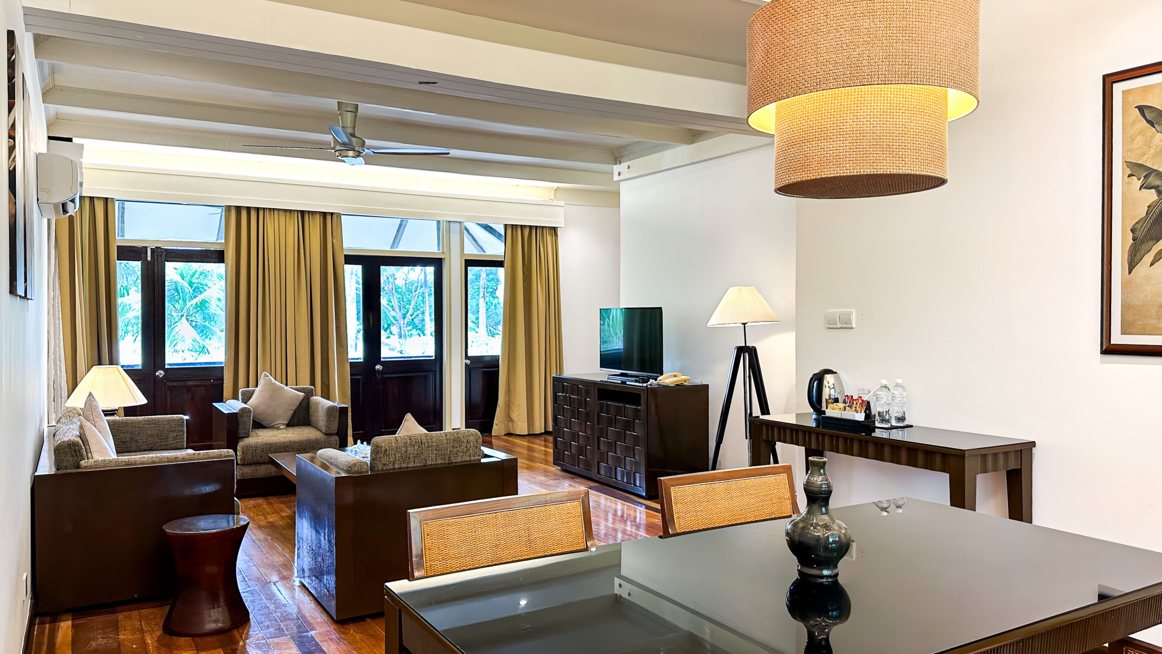 Uma sala de estar espaçosa com área de jantar, janelas grandes com cortinas, um ventilador de teto e uma TV.