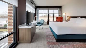 Quarto de hotel com cama, escrivaninha, cadeira, televisão e uma janela grande.