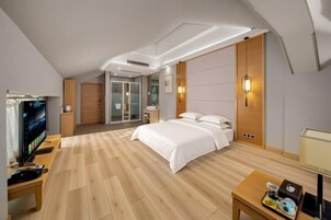 Quarto de hotel moderno com piso de madeira, uma cama, televisão, escrivaninha e banheiro visível por uma porta de vidro.