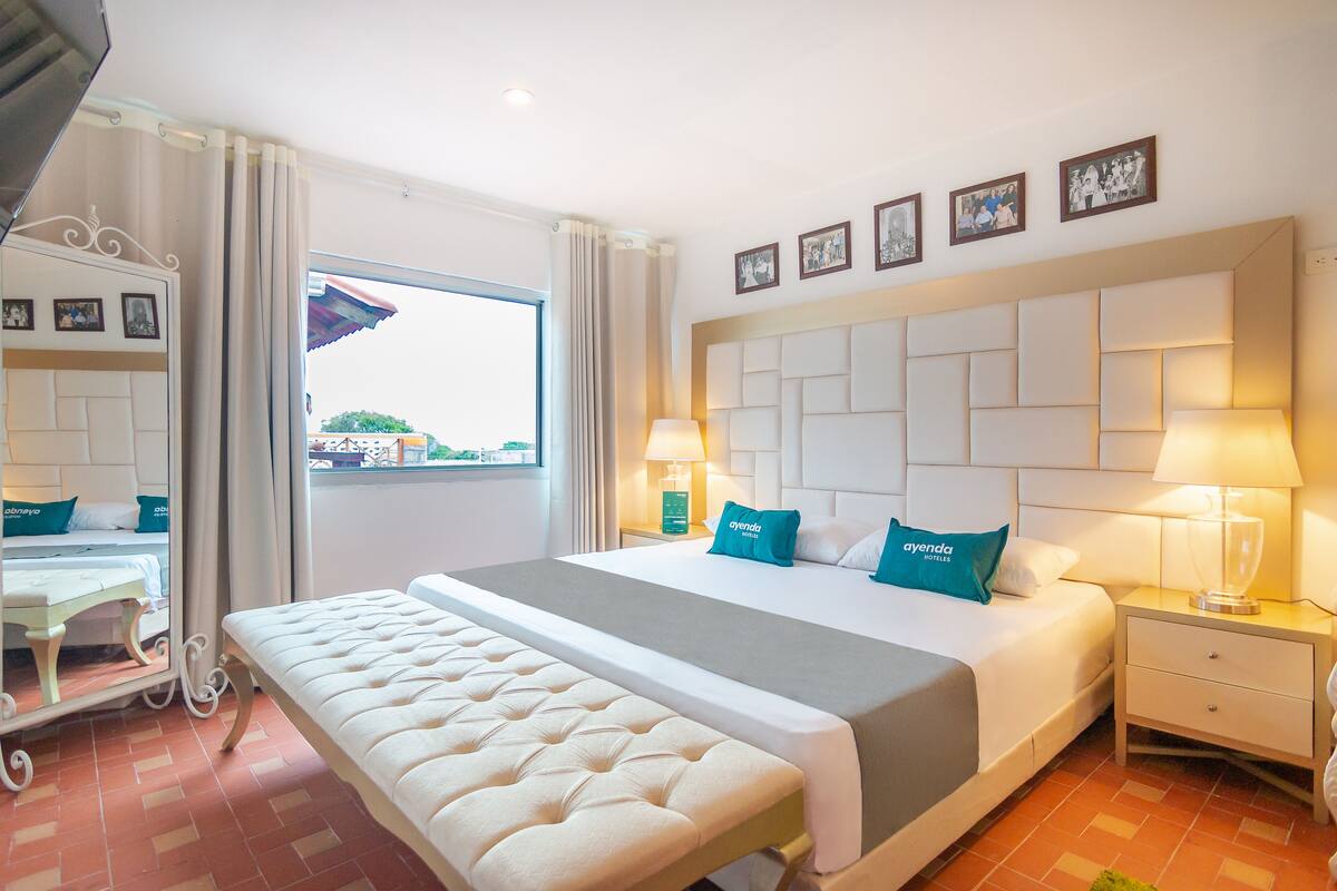 Quarto de hotel com uma cama grande, duas mesas de cabeceira, um banco, um espelho e uma janela com cortinas.