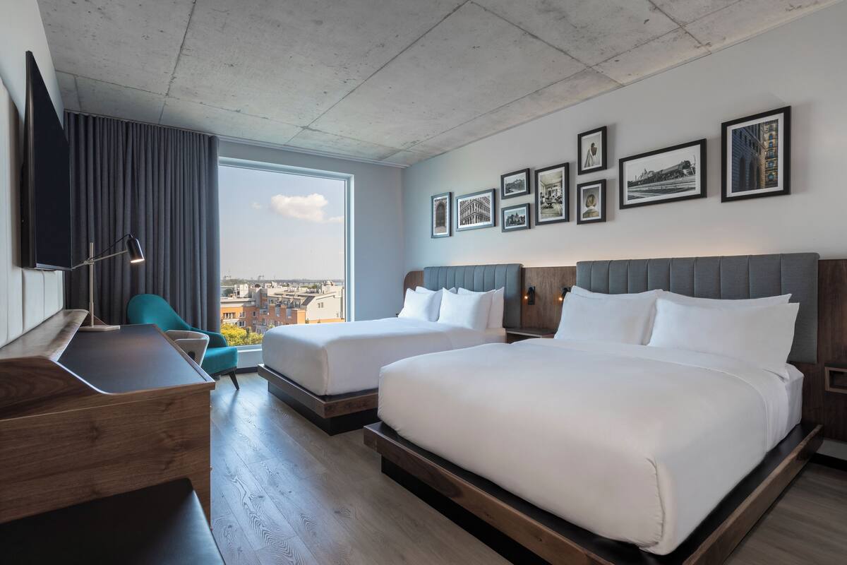 Quarto de hotel com duas camas, uma escrivaninha, uma cadeira e uma janela grande com vista para a cidade.