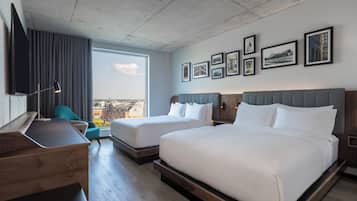 Quarto de hotel com duas camas, uma escrivaninha, uma cadeira e uma janela grande com vista para a cidade.