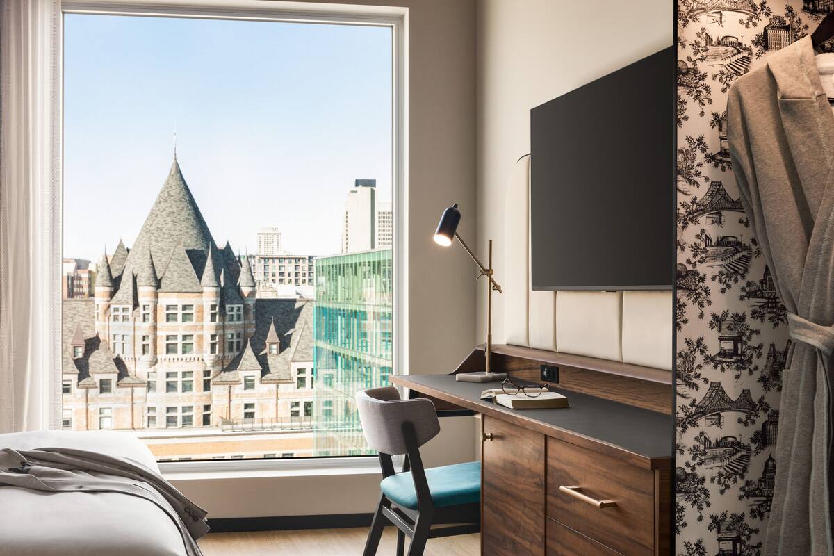 Quarto de hotel com uma grande janela que oferece vista para o horizonte urbano, uma escrivaninha com cadeira, um abajur e uma televisão de tela plana na parede.