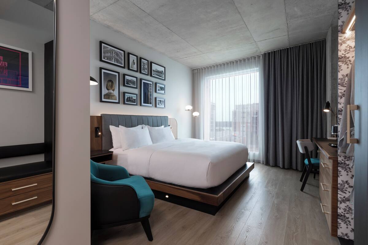 Quarto de hotel moderno com uma cama grande, uma escrivaninha e uma cadeira. Há quadros nas paredes e uma janela com cortinas.