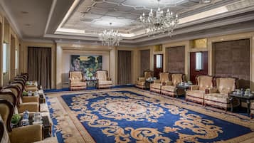 Uma sala de reuniões formal com um grande tapete azul, cadeiras ornamentadas, um candelabro e um quadro na parede.