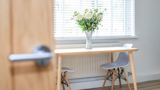 Um quarto com uma mesa de madeira, duas cadeiras cinzas, um vaso branco com flores e uma janela com persianas.