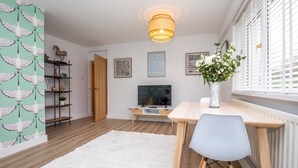 Uma sala de estar moderna com piso de madeira, um tapete branco, uma mesa de jantar com uma cadeira, uma televisão e um vaso com flores.