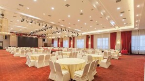Um amplo salão de banquetes com mesas redondas e cadeiras brancas, cortinas vermelhas e um chão coberto por carpete vermelho.