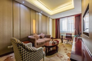 Quarto de hotel com sofá, poltrona e uma pequena mesa. Há um separador de vidro e uma escrivaninha com cadeira.