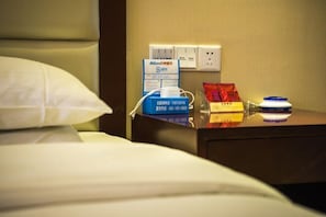 Quarto de hotel com uma cama, uma mesa de cabeceira com régua de tomada, uma caixa azul, um pacote vermelho e um recipiente branco.