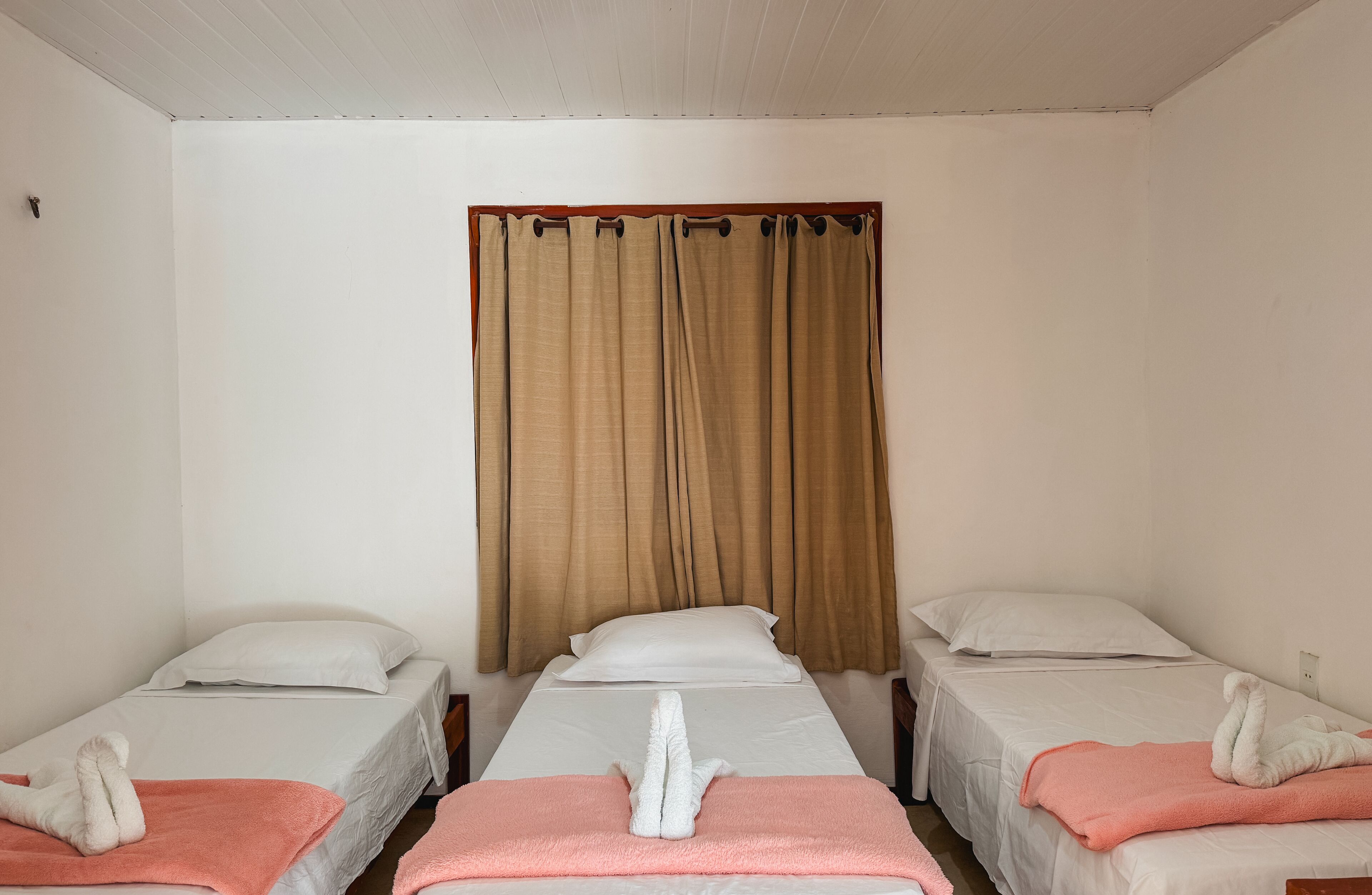 Quarto de hotel com três camas de solteiro, cada uma com uma almofada branca e um cobertor rosa.