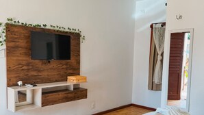 Quarto moderno com suporte de TV de madeira, televisão de tela plana, prateleira de madeira pequena, toalha felpuda grande e espelho com persiana marrom.