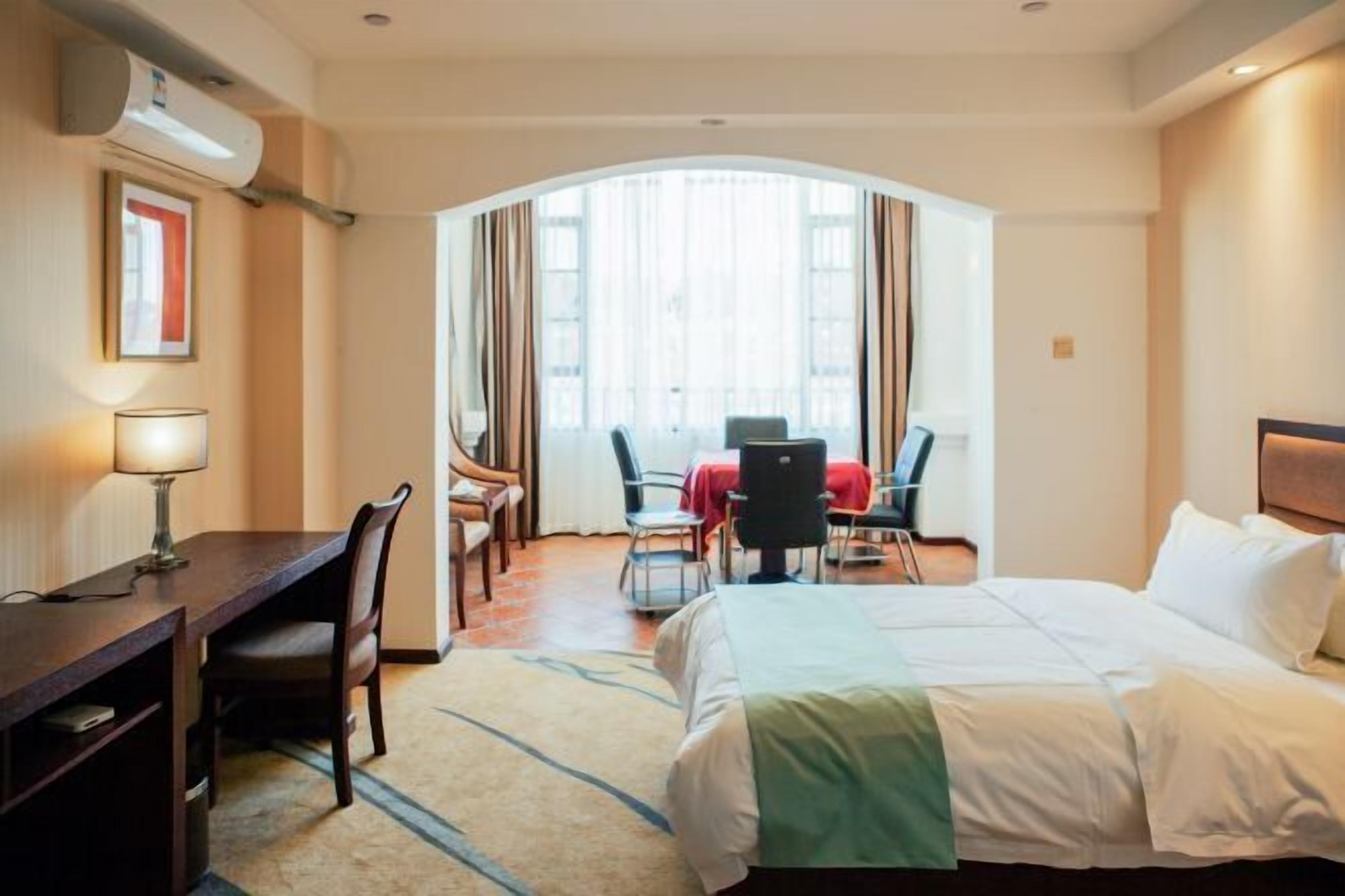 Quarto de hotel com cama, escrivaninha, cadeiras e vista para a rua através de uma janela.