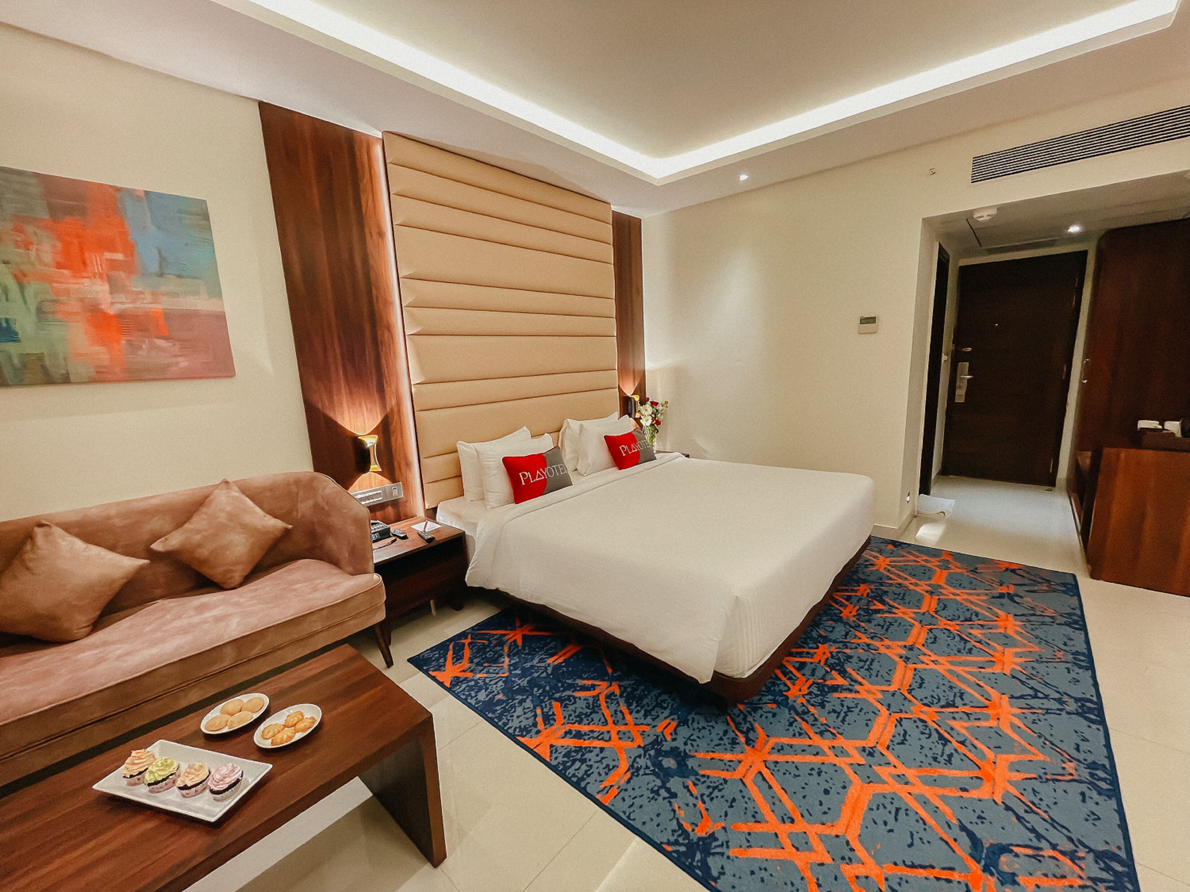 Quarto de hotel com uma cama, um sofá, uma mesa de centro com pastelaria e um tapete colorido.