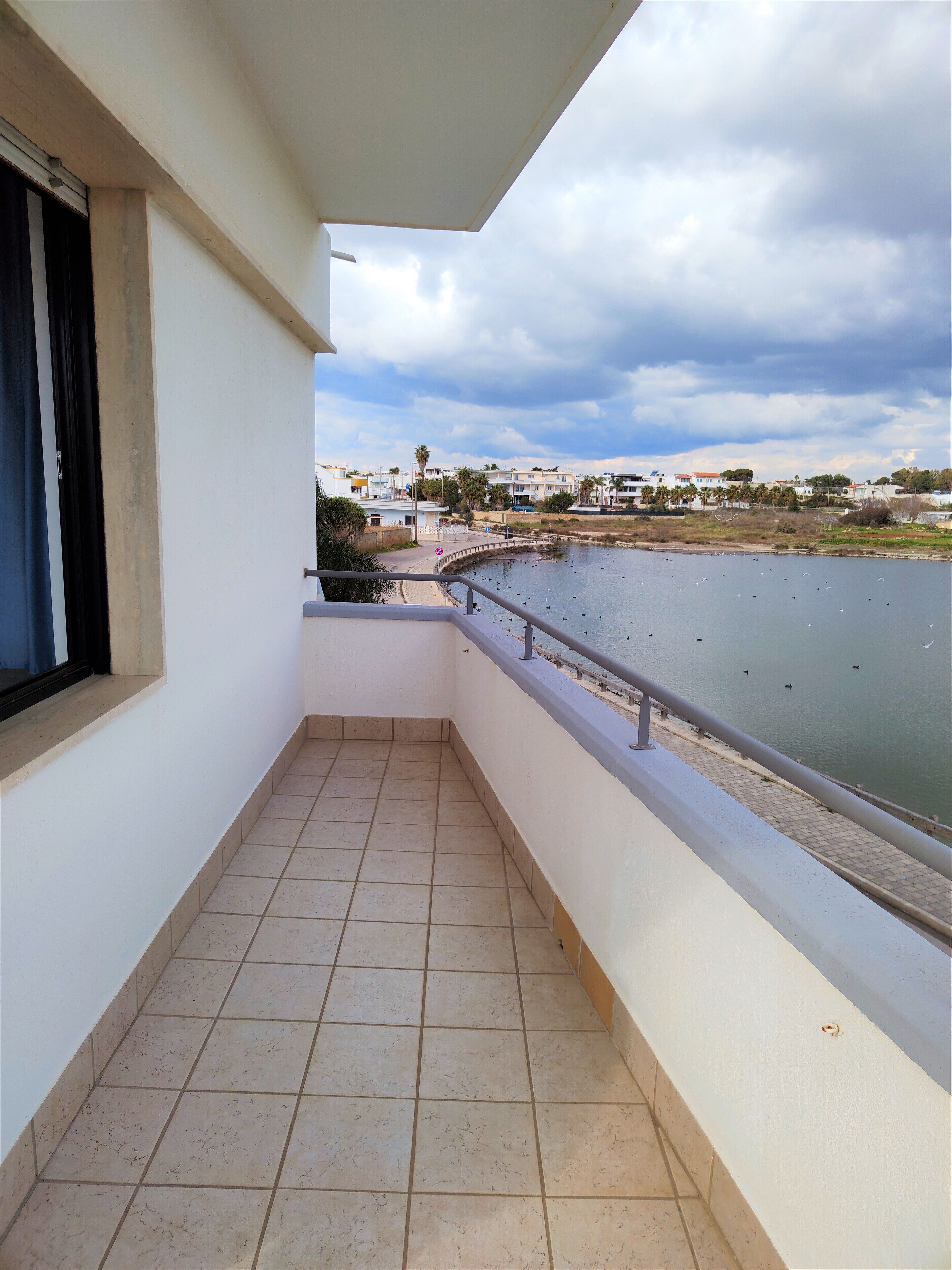 Varanda com chão de azulejos, corrimão e vista para um corpo de água com aves.