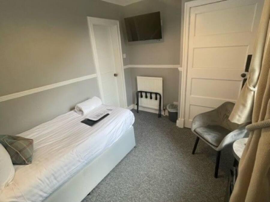 Quarto de hotel com uma cama de solteiro, uma cadeira, televisão fixada na parede e uma porta.