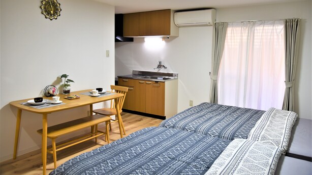 Quarto compacto com cama, pequeno espaço de cozinha, mesa de jantar com cadeiras e janela com cortinas.
