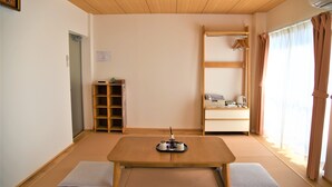 Quarto com piso de tatame, uma mesa de madeira baixa, uma estante com objetos, um guarda-roupa e uma janela com cortinas.