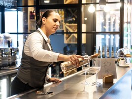 Um bartender servindo uma bebida em um copo em um bar, com um balcão, copos e garrafas ao fundo.