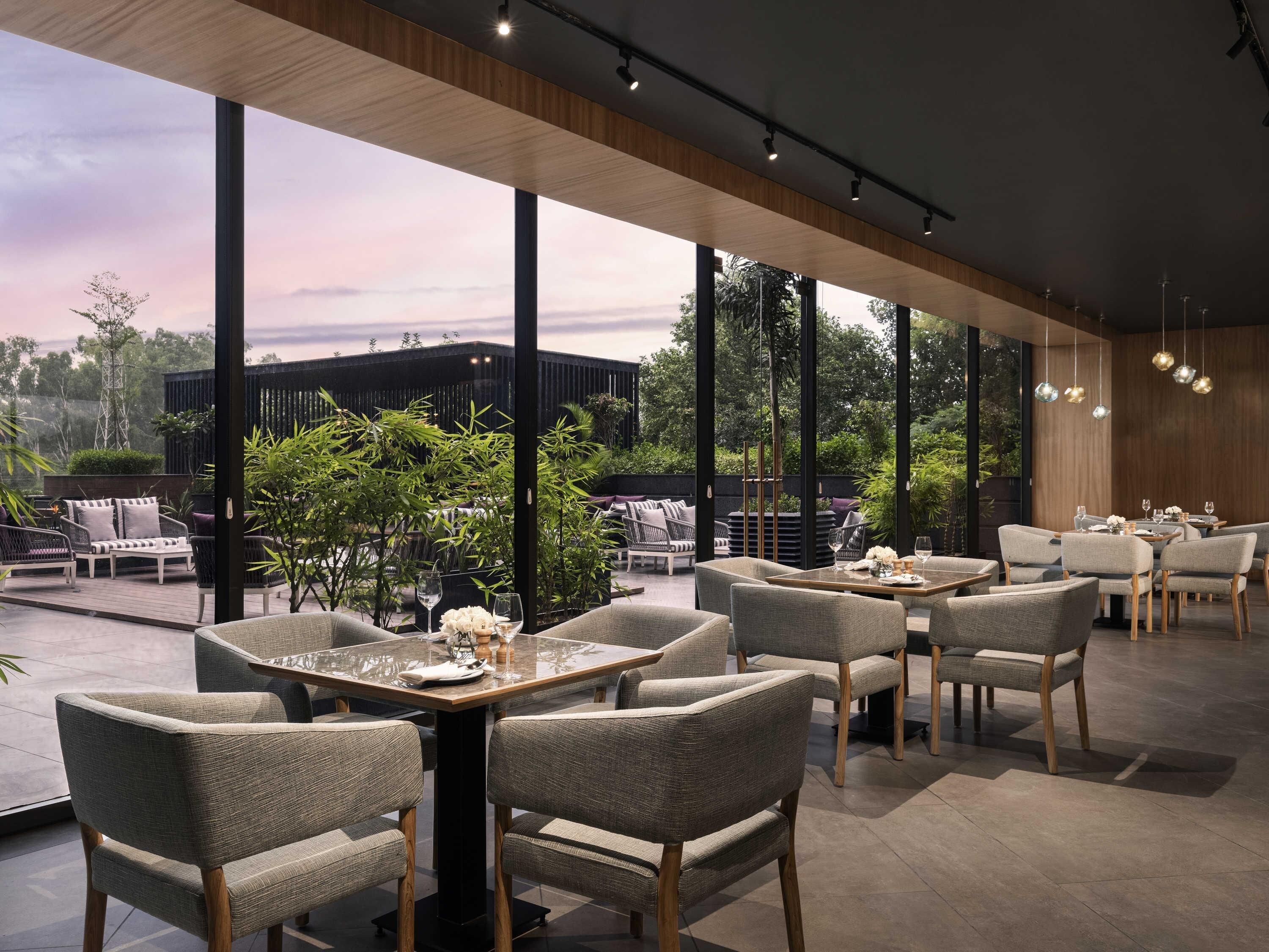Um restaurante moderno com janelas amplas, mesas ao ar livre e vista para o verde.