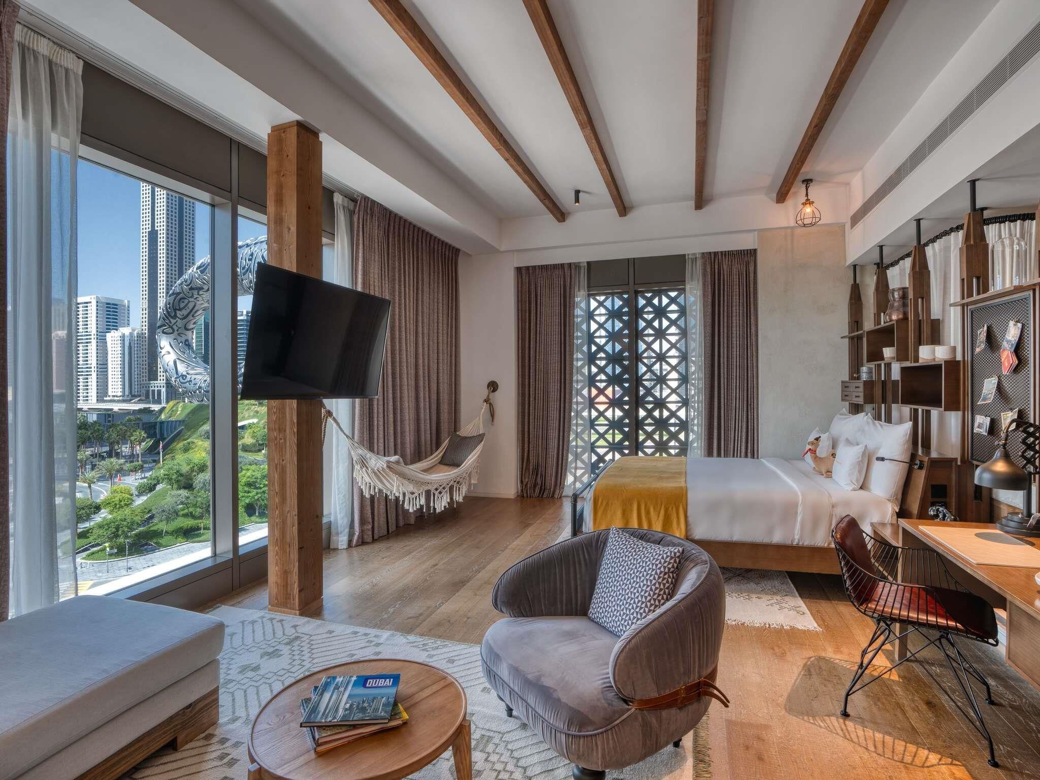 Quarto de hotel moderno com uma cama grande, televisão, rede, sofá e escrivaninha com cadeira.