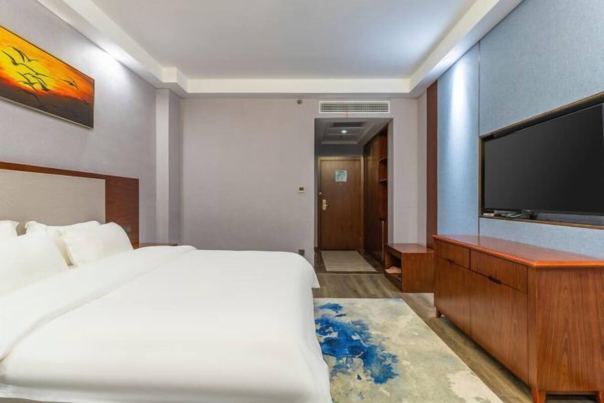 Quarto de hotel com uma cama grande, televisão, aparador de madeira e um quadro na parede.
