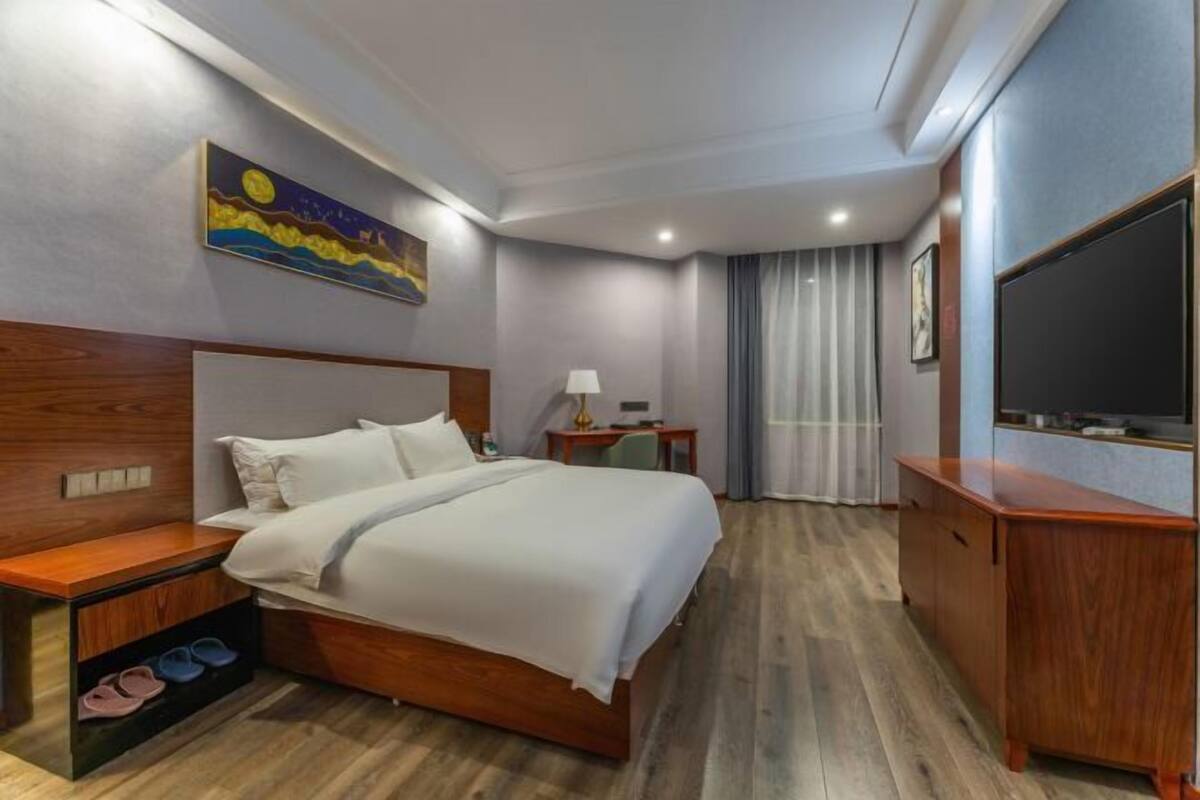 Quarto de hotel com cama de madeira, uma escrivaninha com abajur, televisão e um quadro na parede.