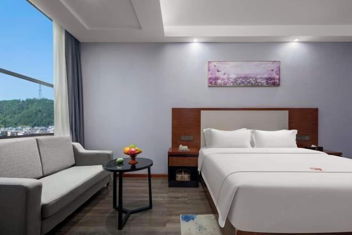 Quarto de hotel com cama, sofá, pequena mesa com abajur, mesa de cabeceira e um quadro na parede.