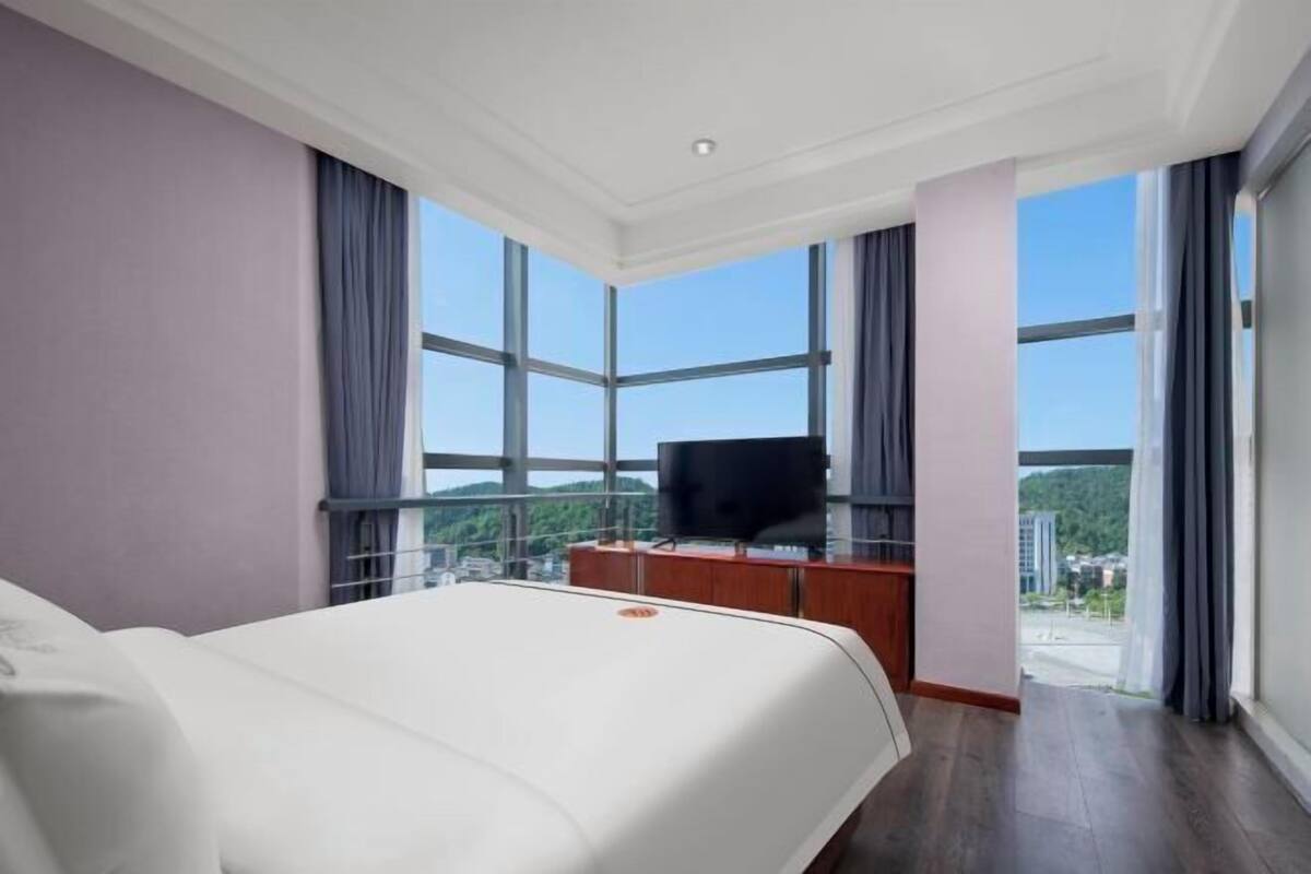 Quarto de hotel com uma cama grande, uma mesa de trabalho com televisão e vista para o horizonte urbano através de amplas janelas.