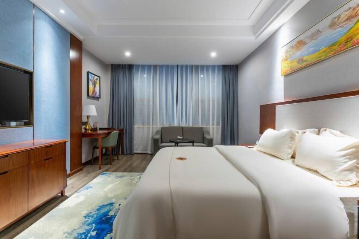 Quarto de hotel com cama, escrivaninha, cadeira, televisão e um quadro na parede.